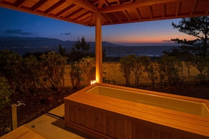 Uma banheira de madeira sob uma pérgola de madeira, com vista para o oceano e montanhas ao anoitecer.