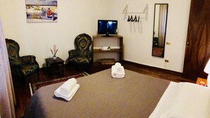 Quarto de hotel com uma cama, duas poltronas, uma televisão, uma mesinha com um vaso e um espelho na parede.