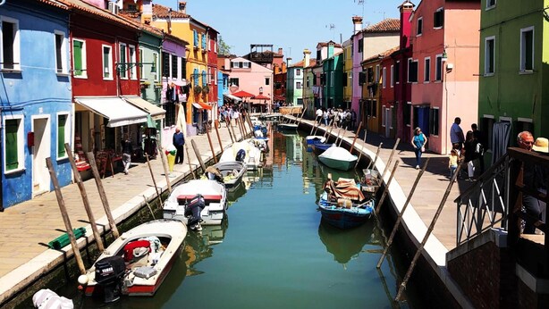 Um canal ladeado por edifícios coloridos, com barcos atracados nas margens e pessoas caminhando nos cais.