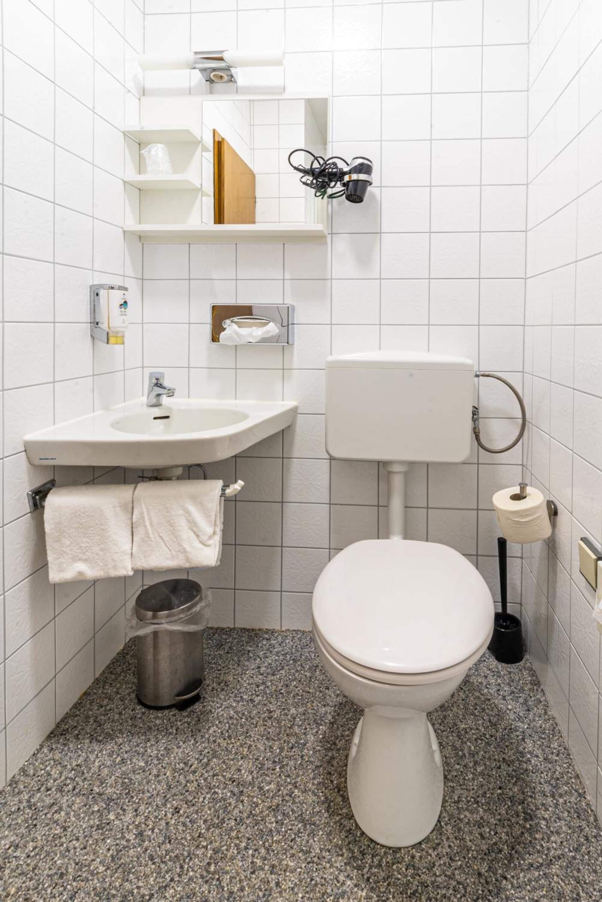 Um banheiro pequeno e limpo, com azulejos brancos, pia branca, vaso sanitário, lixeira de metal e prateleira com artigos de higiene pessoal.