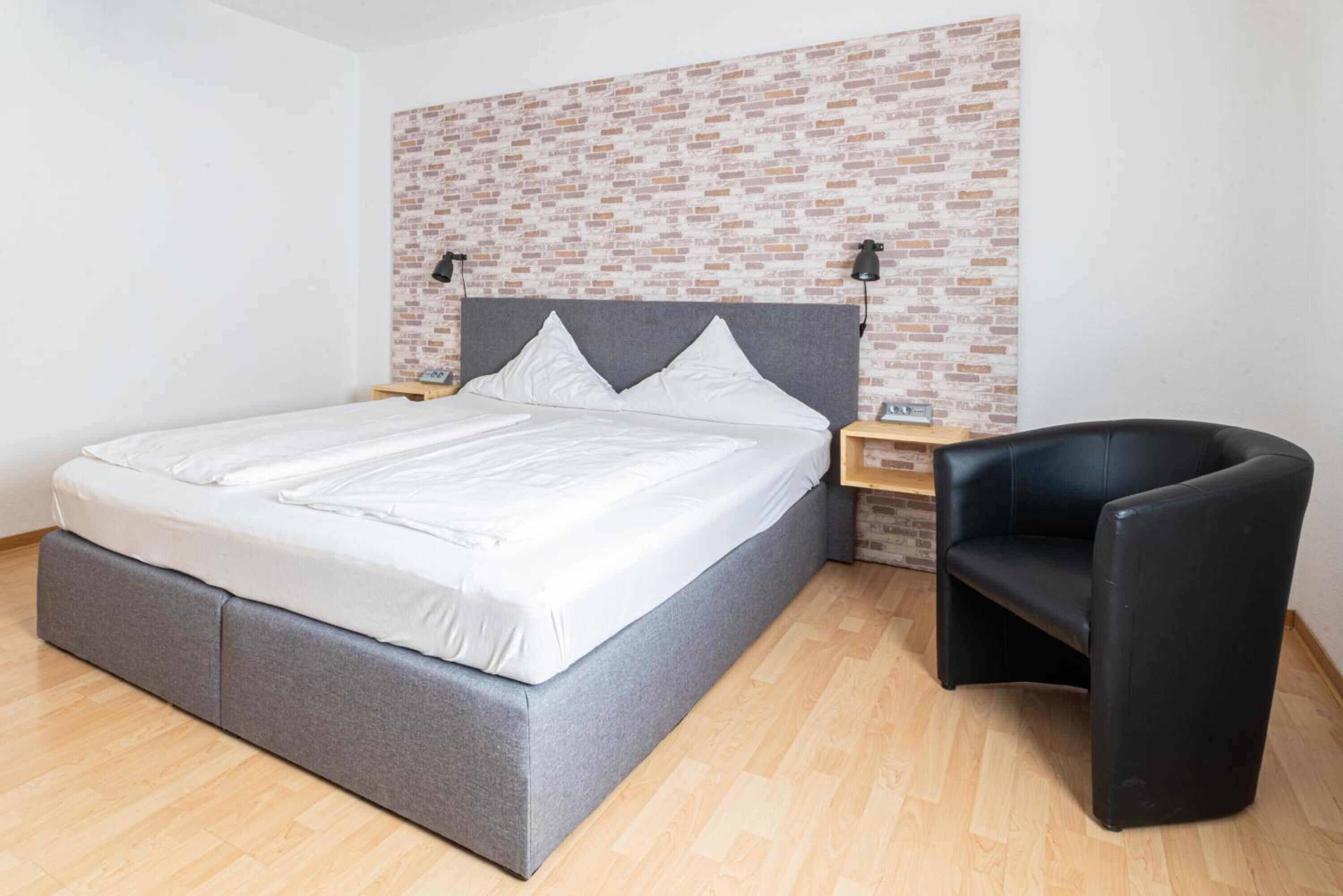 Quarto moderno com cama cinza, cabeceira com padrão de tijolos, poltrona de couro preto e mesa de cabeceira de madeira.
