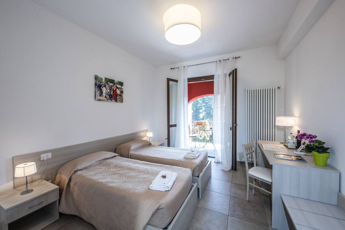 Quarto de hotel com duas camas, uma escrivaninha, uma cadeira, uma mesinha, um abajur, uma planta em vaso e uma janela com cortinas.