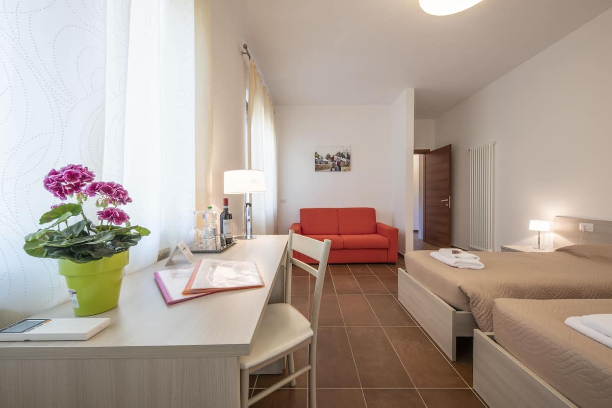 Quarto de hotel com uma mesa, cadeira, duas camas, um sofá vermelho, uma planta em vaso e um quadro na parede.