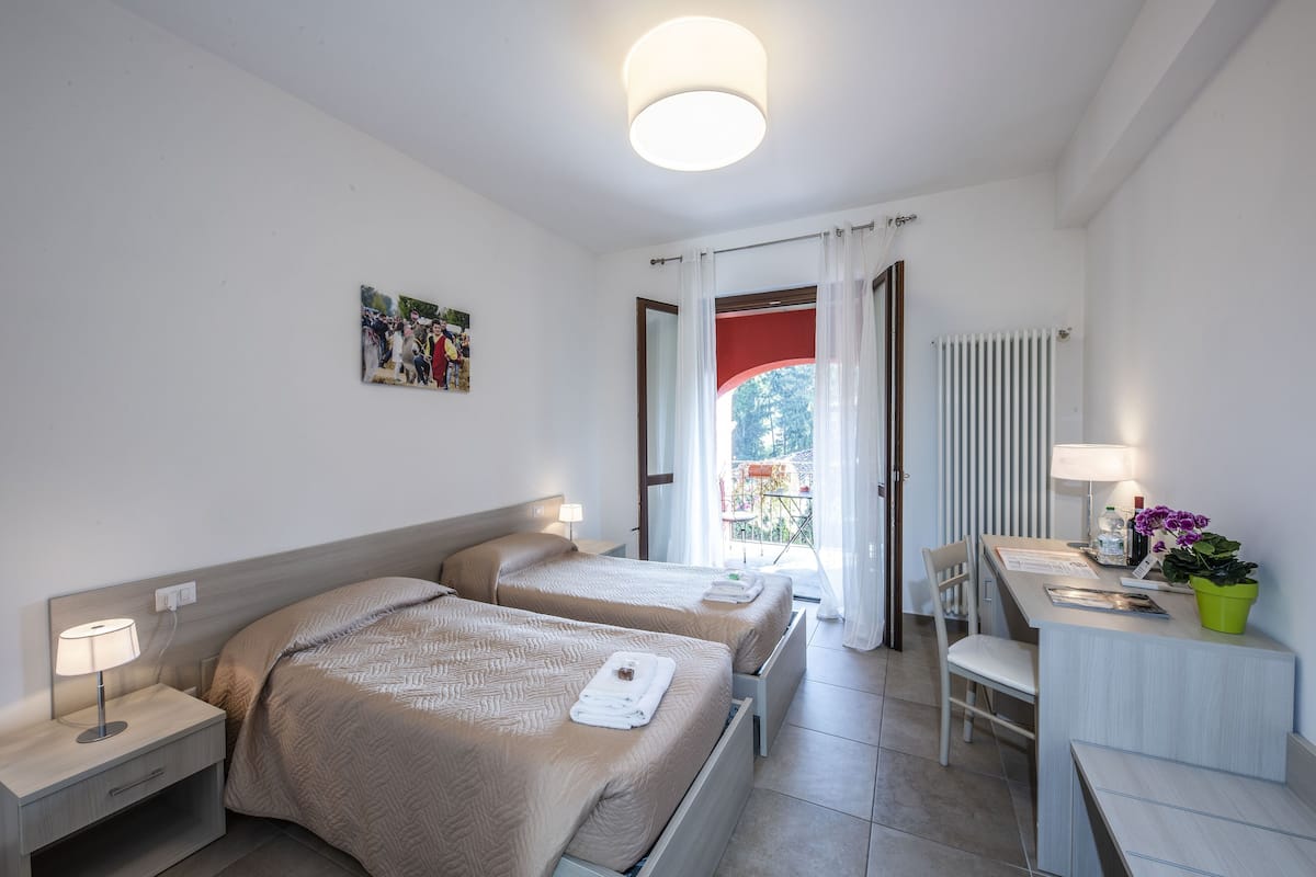 Quarto de hotel com duas camas, uma escrivaninha e uma cadeira. Há uma janela com cortinas, um radiador e uma pequena mesa com abajur e planta.