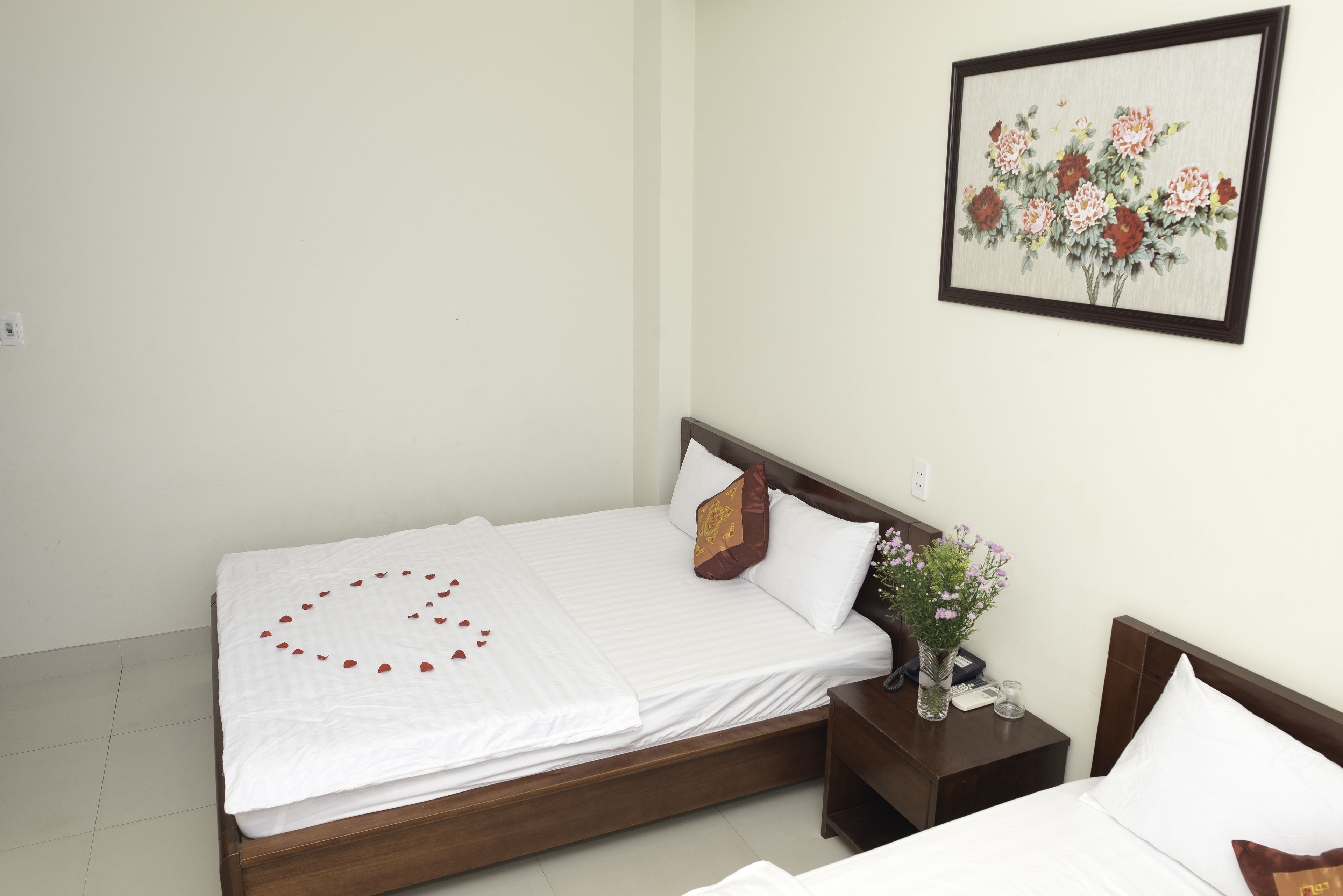 Uma cama bem arrumada com um arranjo de pétalas de rosa em formato de coração, uma mesa de cabeceira com um vaso de flores e uma pintura floral emoldurada na parede.