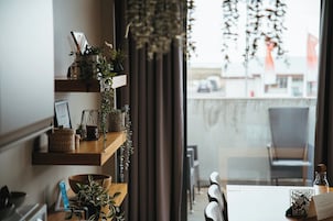 Um interior moderno com uma estante de madeira exibindo objetos decorativos e plantas, uma mesa de jantar com cadeiras, e vista para o exterior através de grandes janelas.