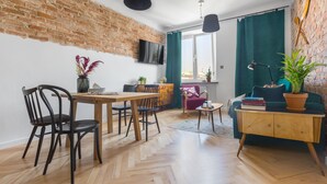 Uma sala de estar moderna com piso de madeira em padrão herringbone, parede de tijolos, sofá verde, mesa de jantar de madeira com cadeiras e uma TV fixada na parede.