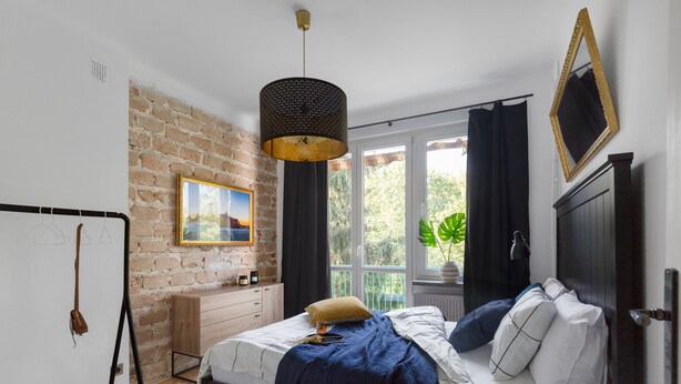 Um quarto com cama, escrivaninha, luminária pendente, janela com cortinas e parede de tijolos.