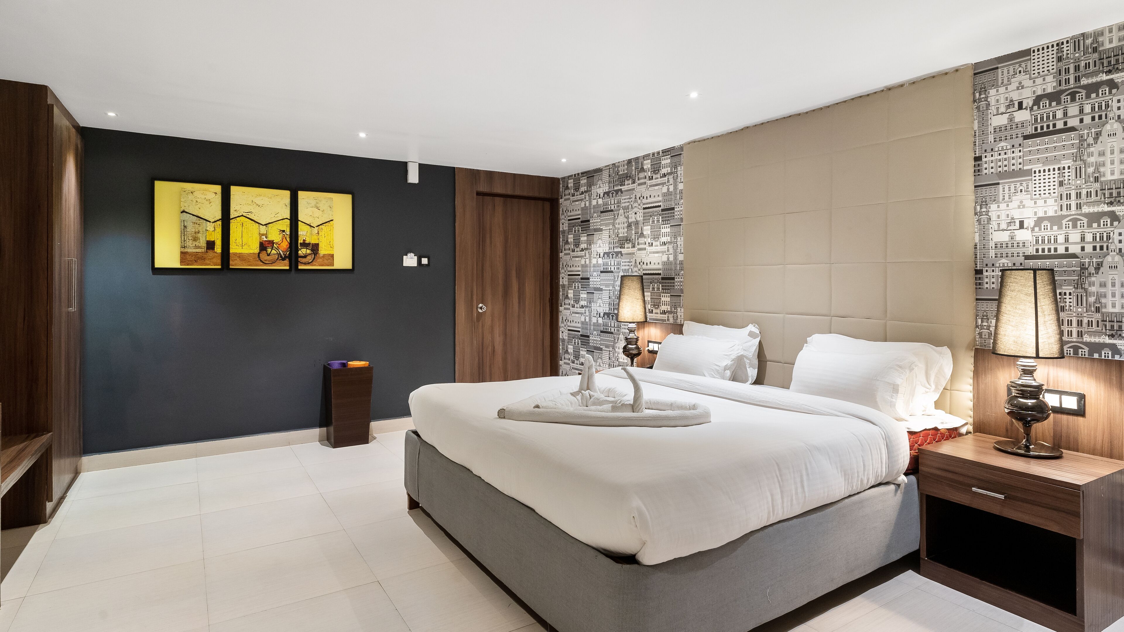 Quarto de hotel moderno com uma cama grande, um guarda-roupa de madeira, uma pequena mesa de cabeceira com abajur e um papel de parede com vista para a cidade.