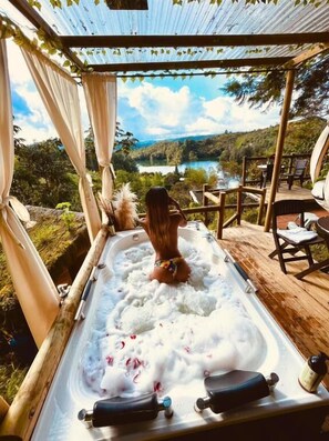 Uma banheira de hidromassagem com uma pessoa sentada no interior, cercada por um deck de madeira e uma vista panorâmica de um lago e colinas.