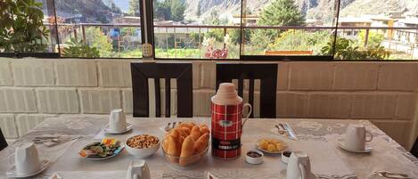 Mesa posta para o café da manhã, com uma garrafa térmica, pão e diversos acompanhamentos, com vista para uma paisagem montanhosa.