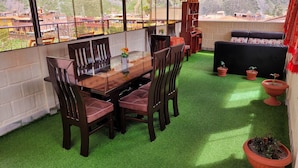Uma área de refeições com mesa e cadeiras de madeira, uma estante de livros e plantas em vasos sobre um piso de grama sintética verde.