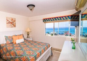 Quarto com cama, mesa de cabeceira, luminária, janela com vista para o mar e uma pequena mesa com um vaso.
