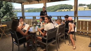 Um grupo de pessoas sentadas em mesas num terraço ao ar livre, com vista para um lago e colinas.