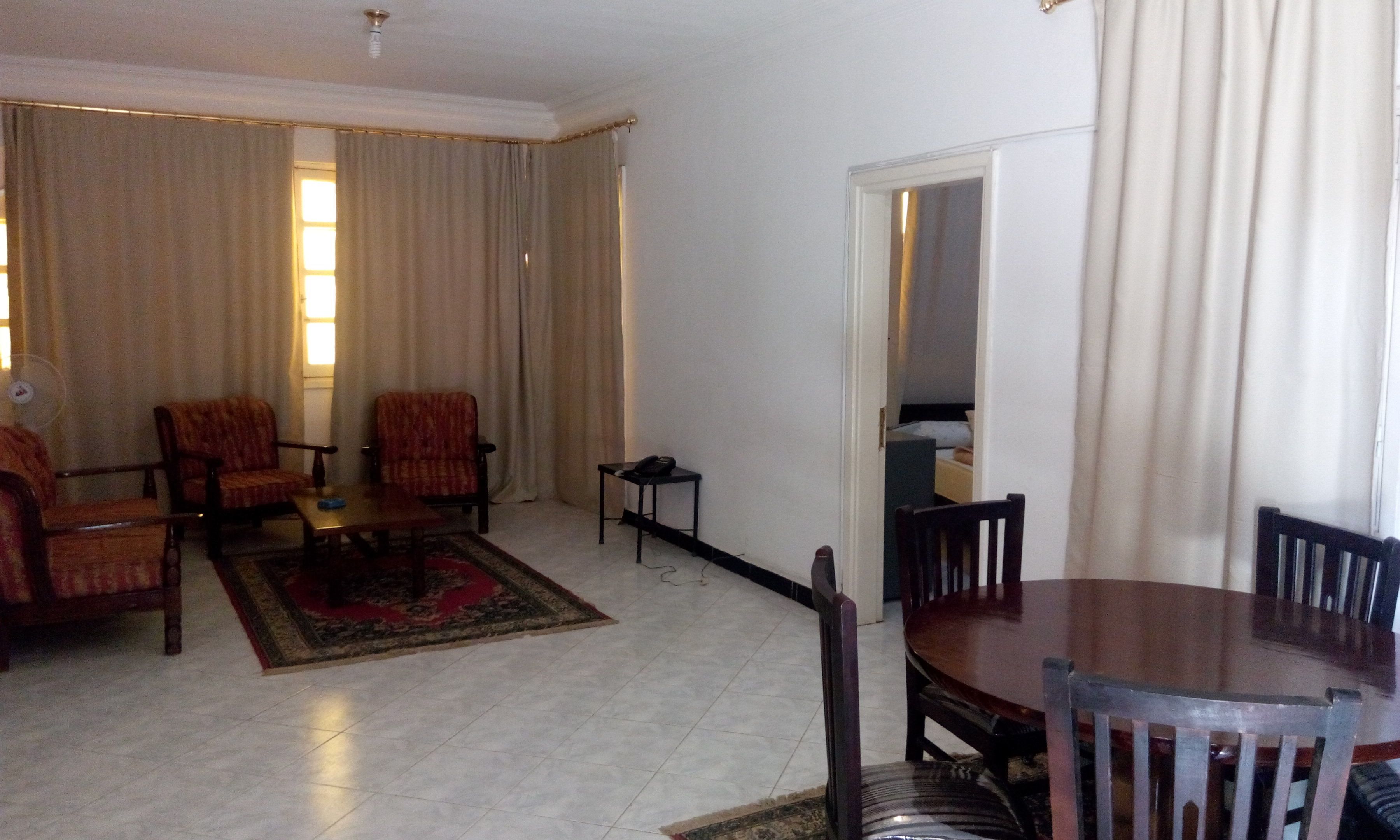Uma sala de estar com uma mesa de jantar, duas poltronas, um sofá e uma mesa de centro. Há uma porta que dá para outro cômodo.