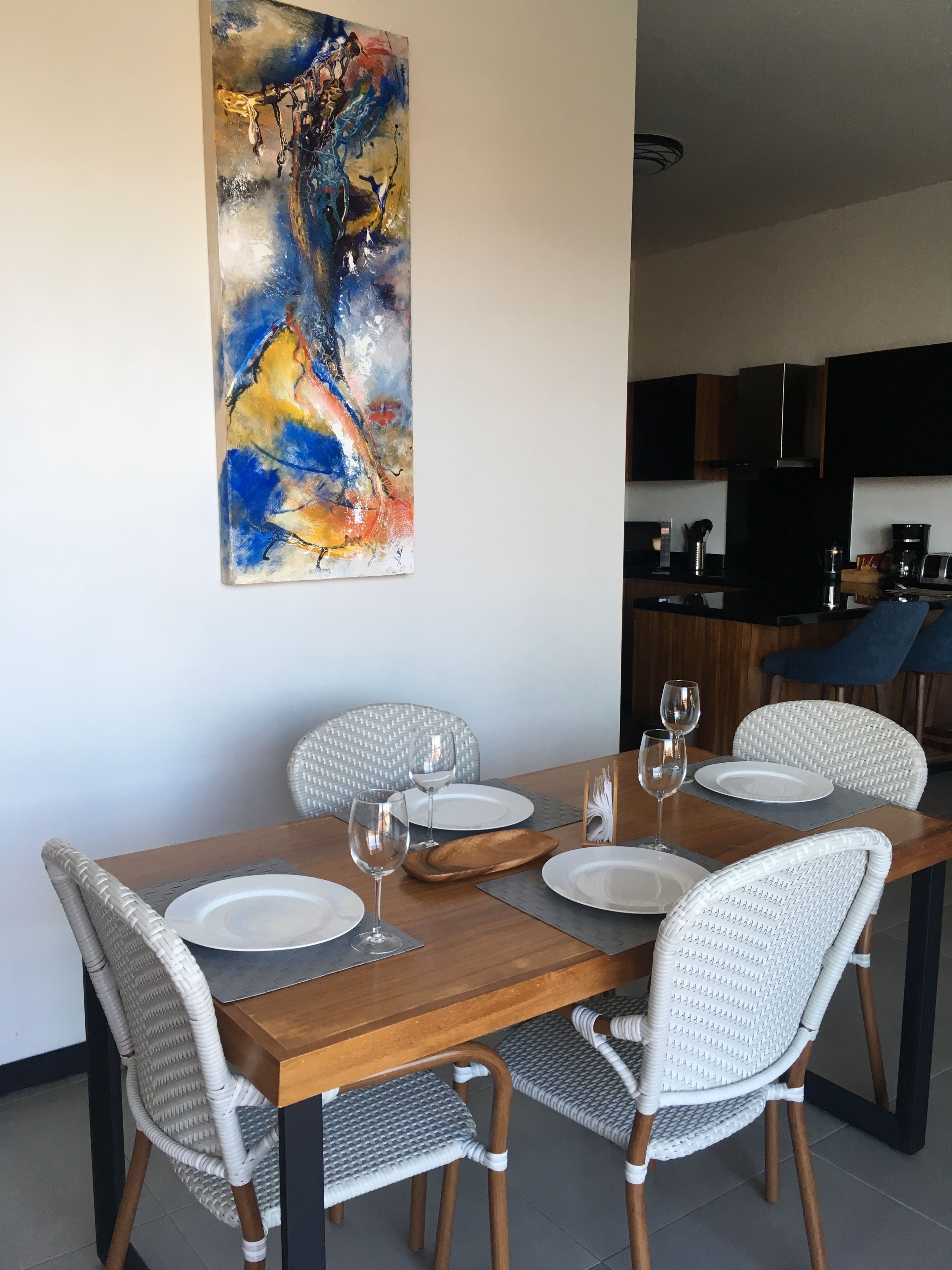 Uma sala de jantar com uma mesa de madeira, cadeiras brancas trançadas e um grande quadro abstrato na parede.
