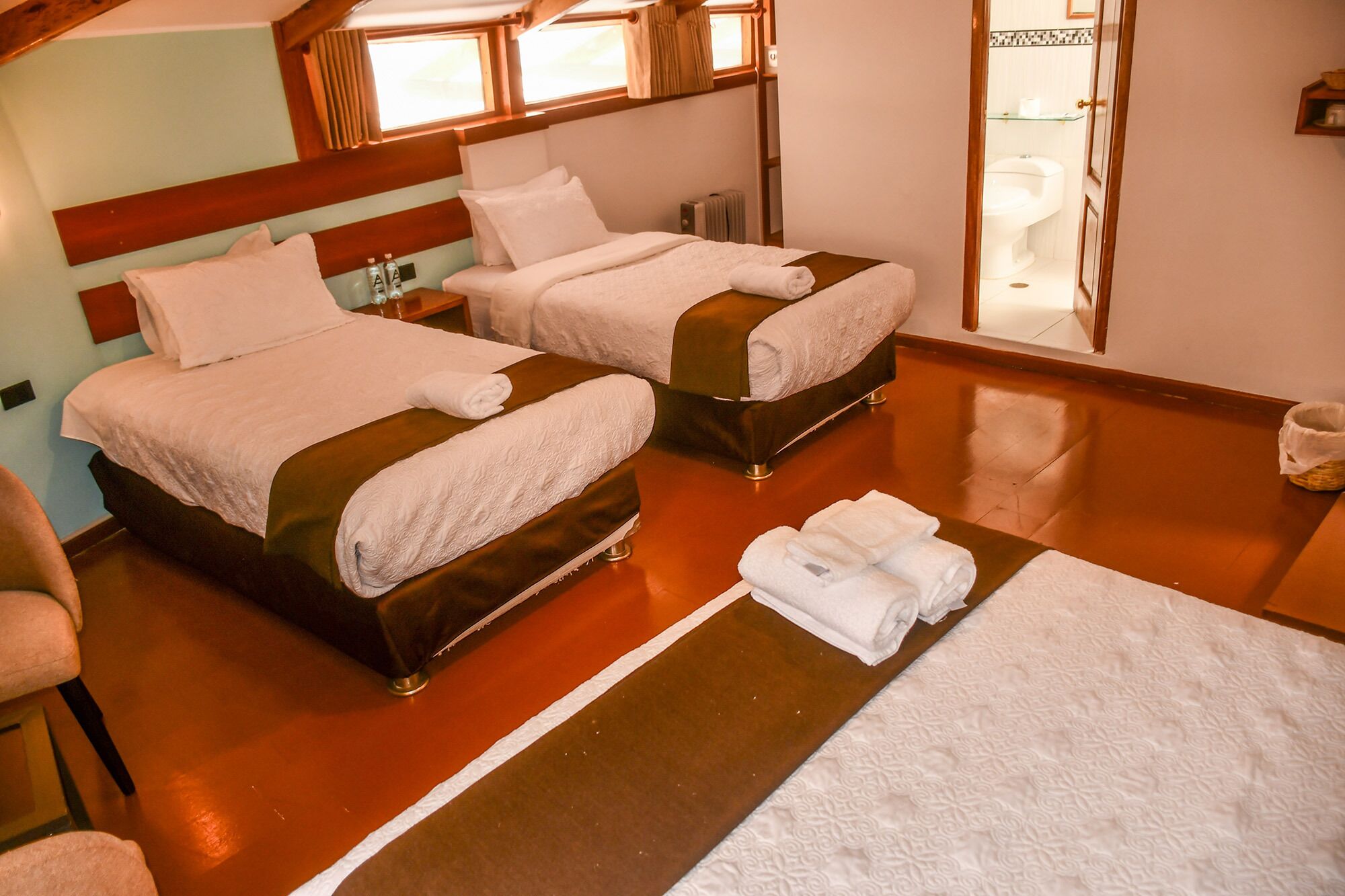 Quarto de hotel com duas camas, cada uma com cabeceira de madeira, uma mesinha com uma garrafa e uma cadeira.