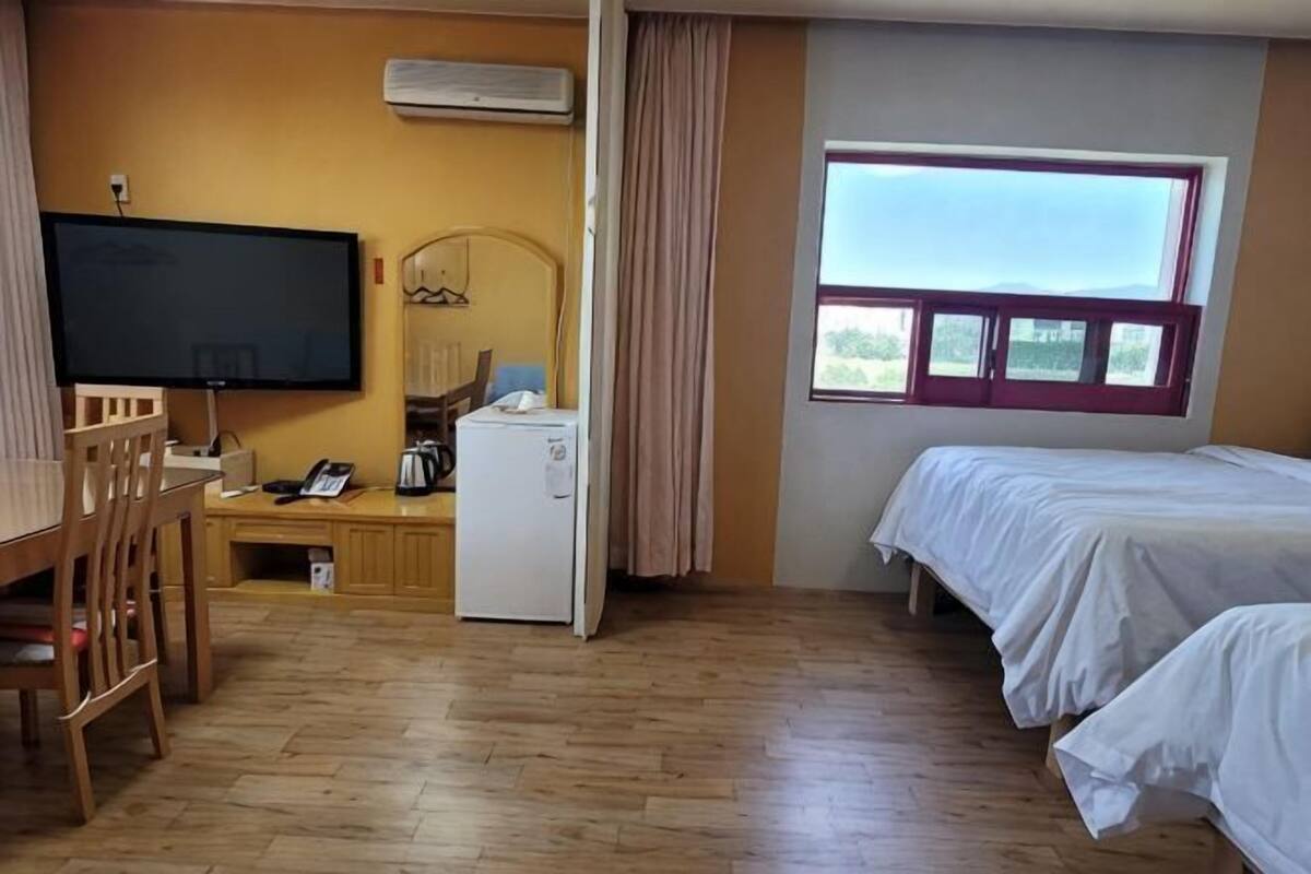 Quarto de hotel com cama, televisão, escrivaninha com cadeira, frigorífico e janela com cortinas.