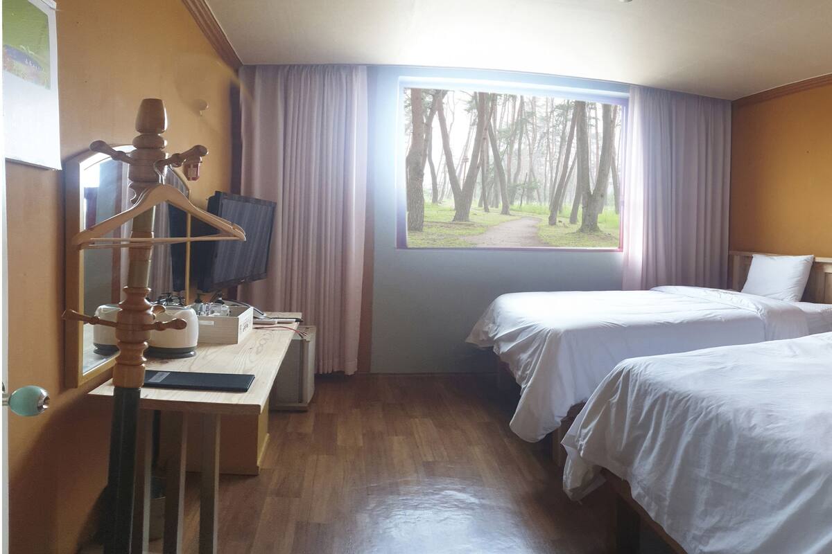 Quarto de hotel com duas camas, uma escrivaninha com computador, um cabideiro de madeira e uma janela com vista para a floresta.