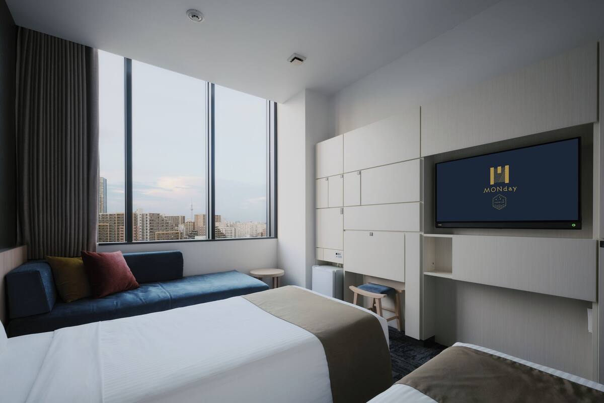Quarto de hotel moderno com uma cama grande, um sofá, uma TV fixada na parede e vista para a cidade pelas janelas.
