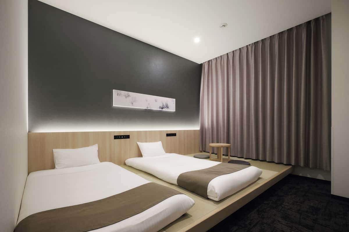 Quarto de hotel com duas camas, uma mesinha, uma luminária na parede e uma janela com cortinas.