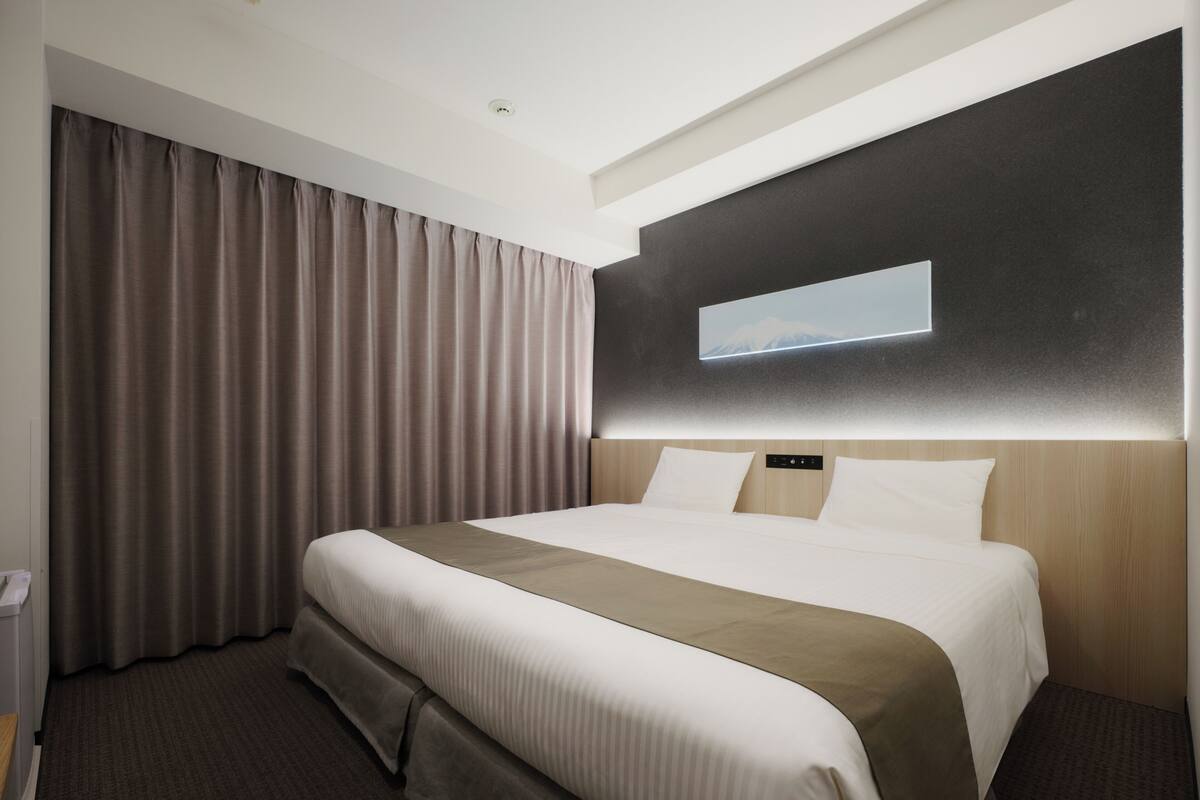 Quarto de hotel com uma cama grande, cabeceira com TV embutida, janela ampla com cortinas e piso coberto por carpete.