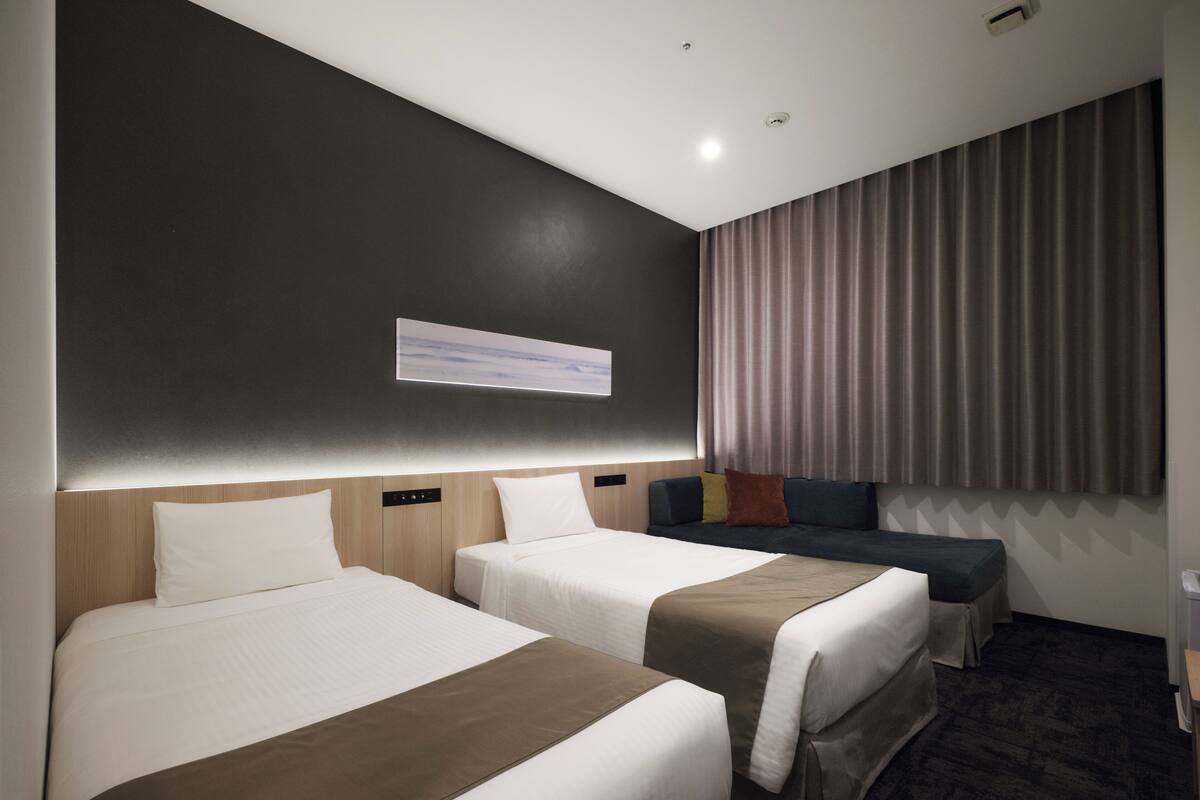 Quarto de hotel com duas camas, um sofá, uma janela grande com cortinas e um candeeiro fixado na parede.