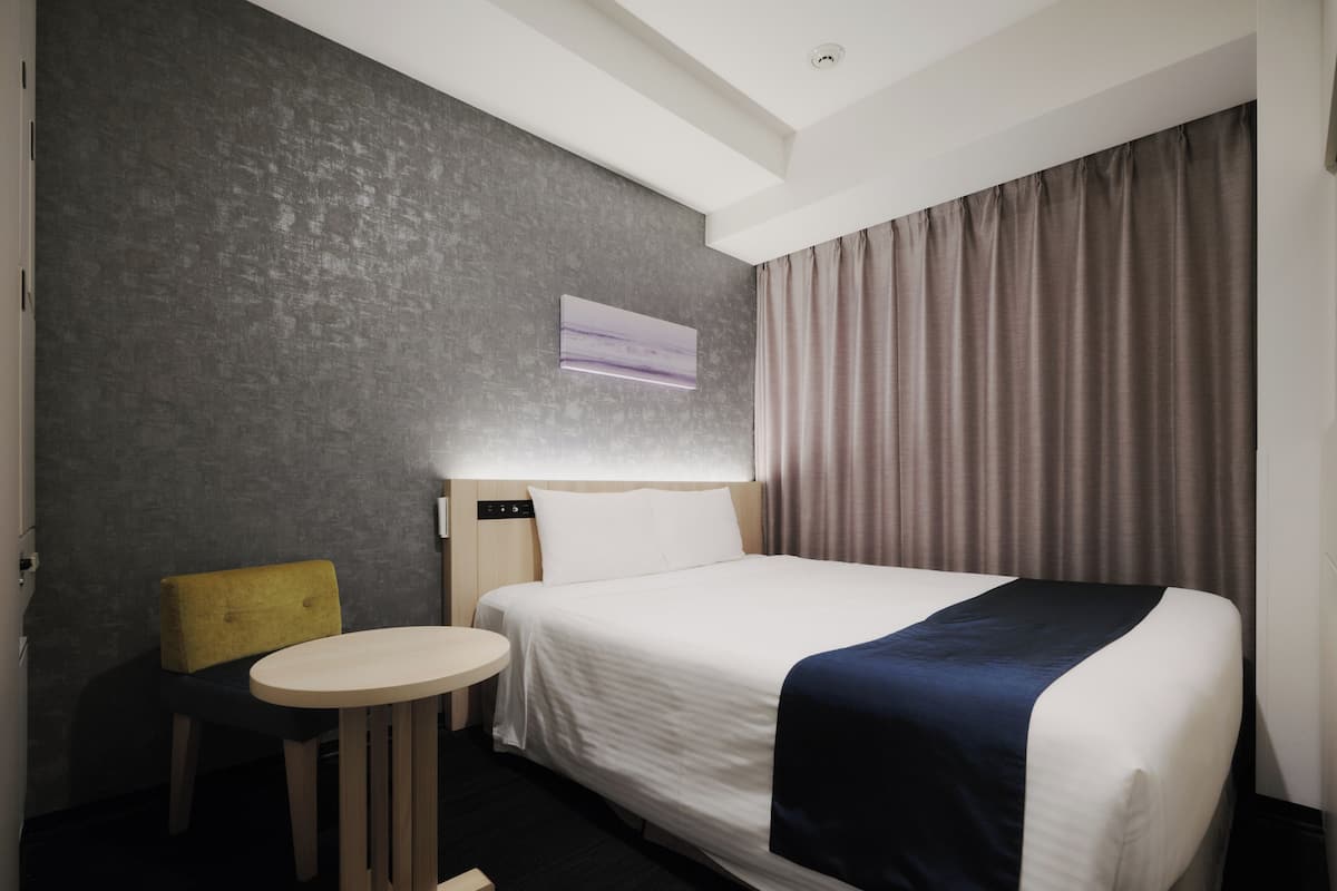 Quarto de hotel com uma cama, uma pequena mesa redonda, uma cadeira e uma parede cinza com textura.