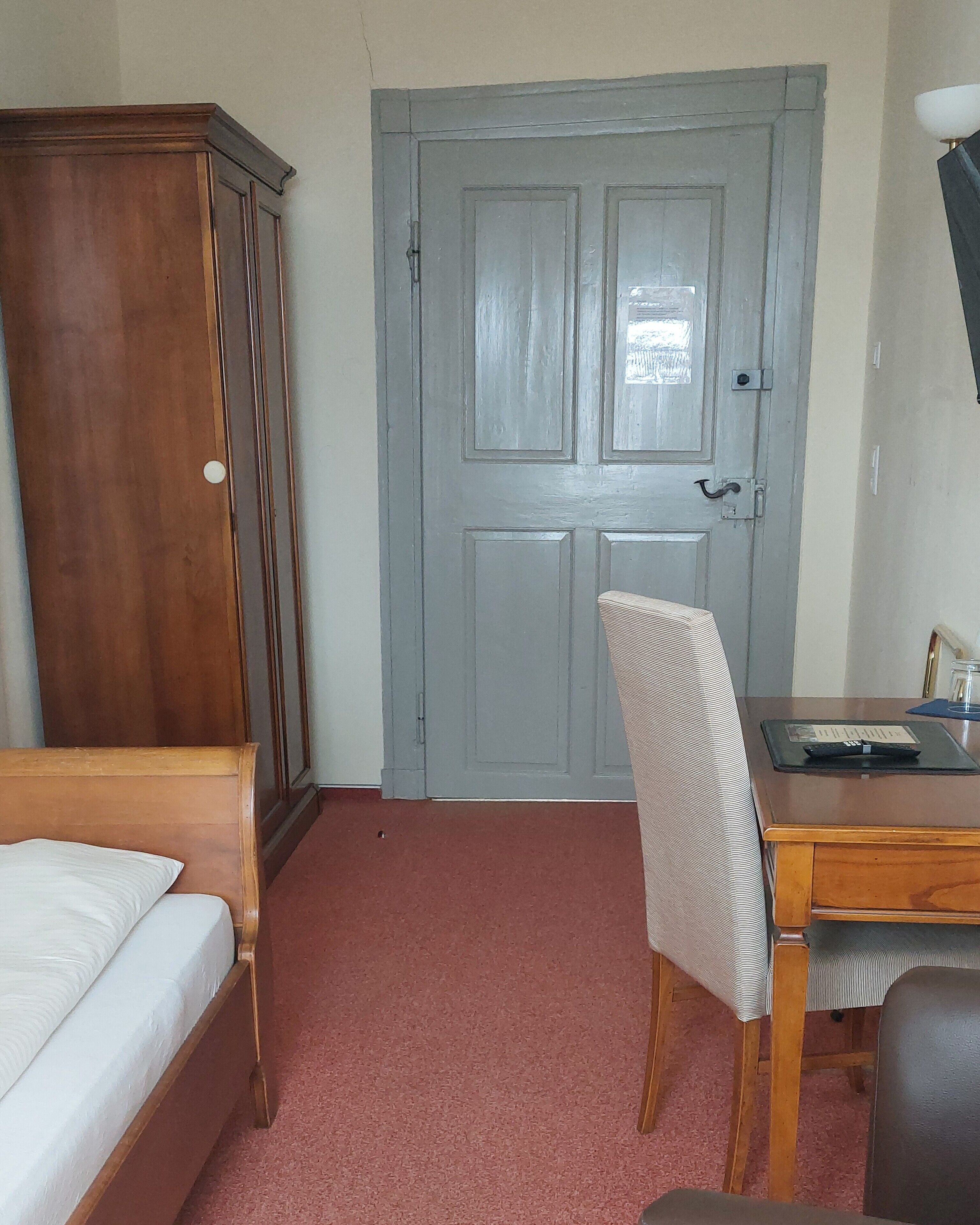 Quarto de hotel com cama de madeira, escrivaninha com cadeira, guarda-roupa e porta cinza.