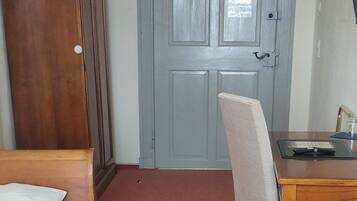 Quarto de hotel com cama de madeira, escrivaninha com cadeira, guarda-roupa e porta cinza.