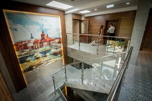 Um interior com vários níveis, corrimão de vidro, uma pintura grande de um edifício histórico e uma porta de madeira.