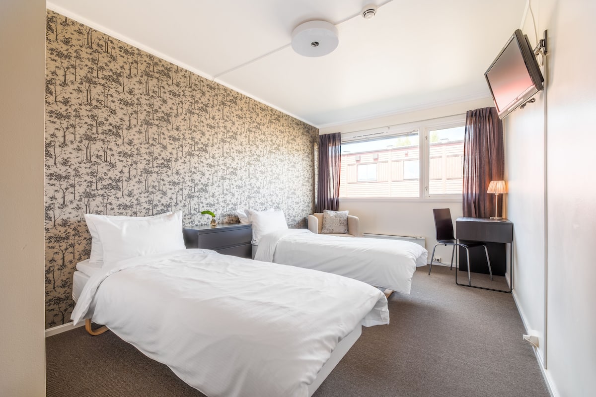 Quarto de hotel com duas camas, uma escrivaninha, uma cadeira, uma televisão e uma janela com cortinas.
