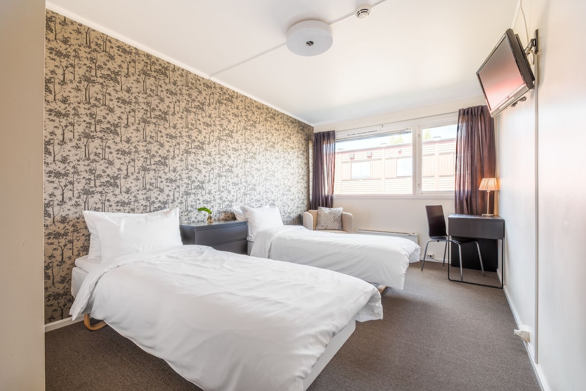 Quarto de hotel com duas camas, uma escrivaninha, uma cadeira, uma televisão e uma janela com cortinas.