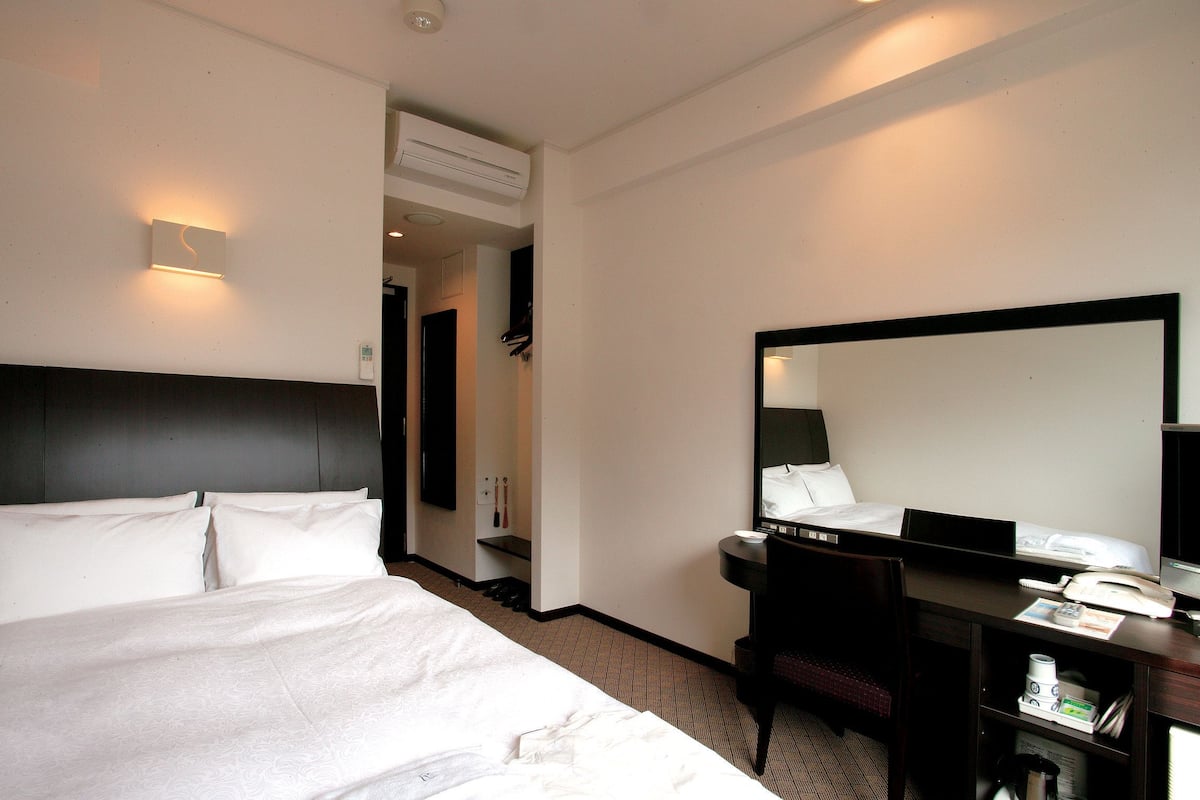 Quarto de hotel com uma cama grande, uma mesa com cadeira, um espelho e um ar condicionado fixado na parede.