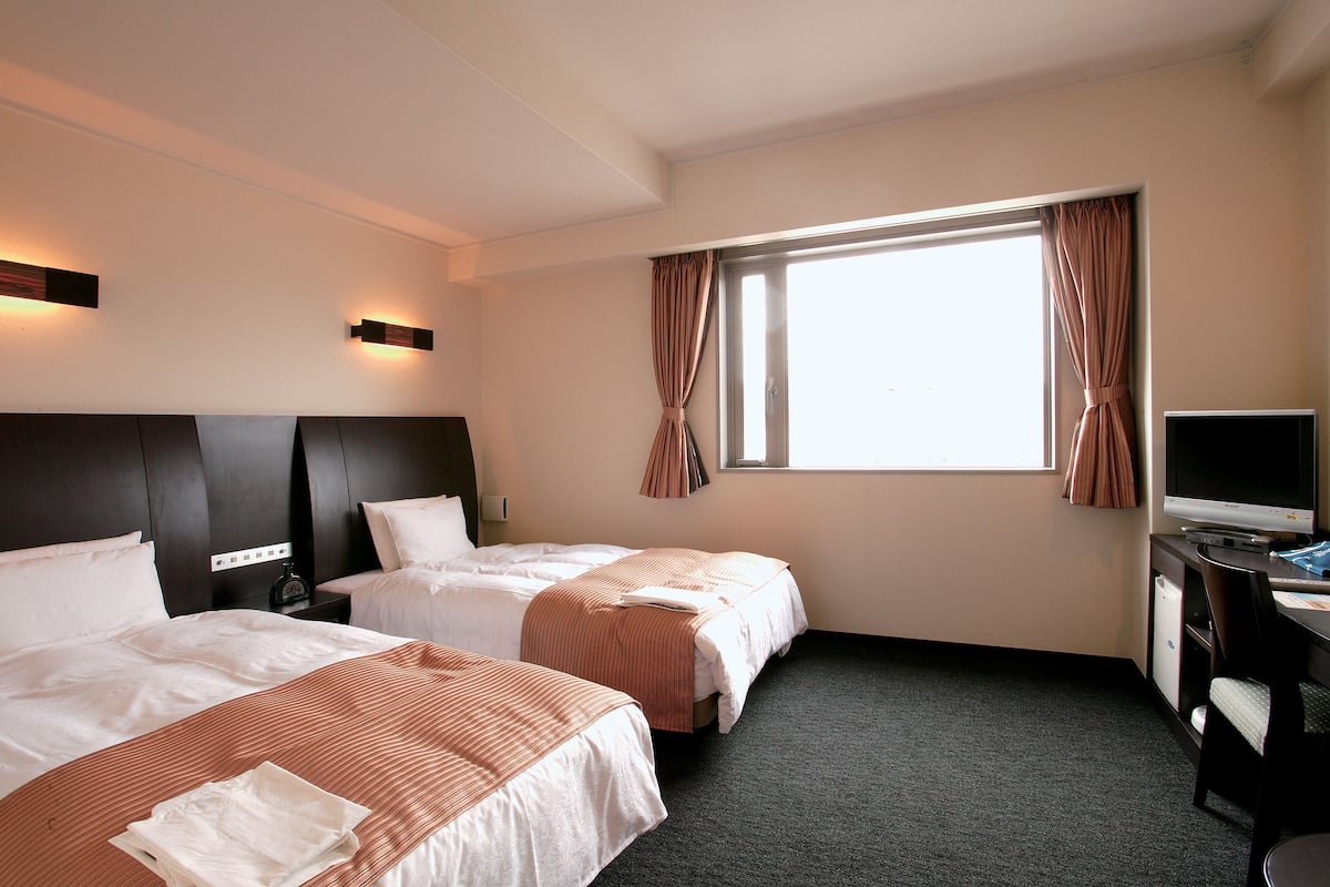 Quarto de hotel com duas camas, uma escrivaninha, uma cadeira, televisão e janela com cortinas.