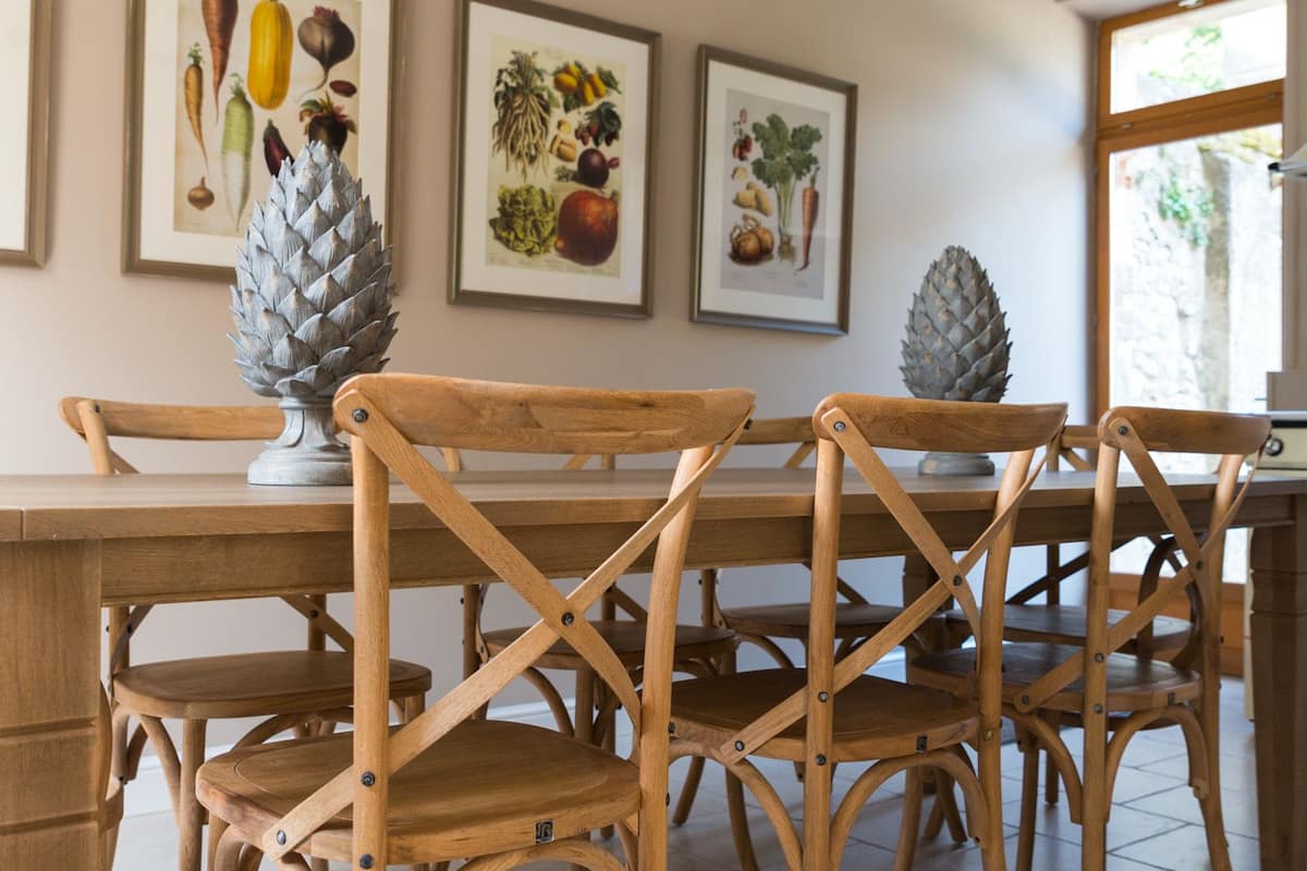 Uma área de refeições com cadeiras de madeira e uma mesa, quadros botânicos nas paredes e um lustre.