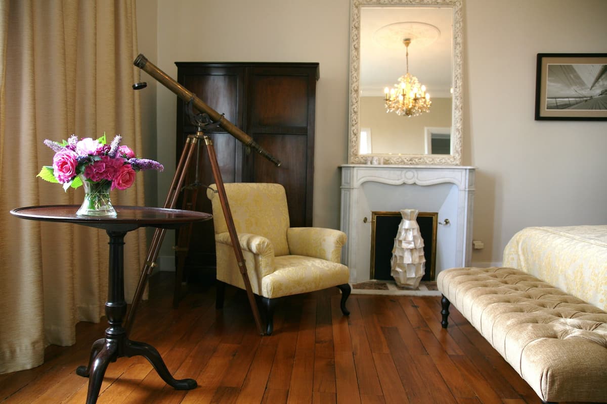 Um quarto com piso de madeira, uma mesa redonda com um vaso de flores, uma poltrona amarela, uma lareira branca, uma cama e um espelho grande com um candelabro.