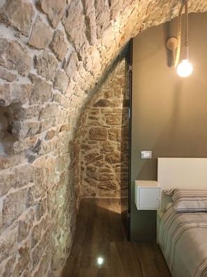 Um quarto com parede de pedra, piso de madeira, uma cama com roupa de cama listrada e uma mesinha de cabeceira.