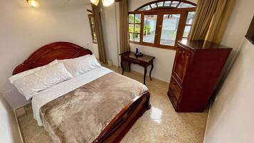 Quarto com cama de madeira, escrivaninha, guarda-roupa e ventoinha no teto.