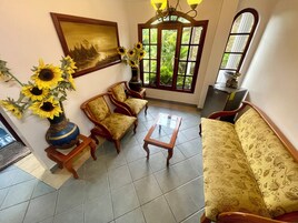 Uma sala de estar com sofá, duas poltronas, uma mesa de centro, um quadro, um vaso com girassóis e uma janela com vista para o verde.