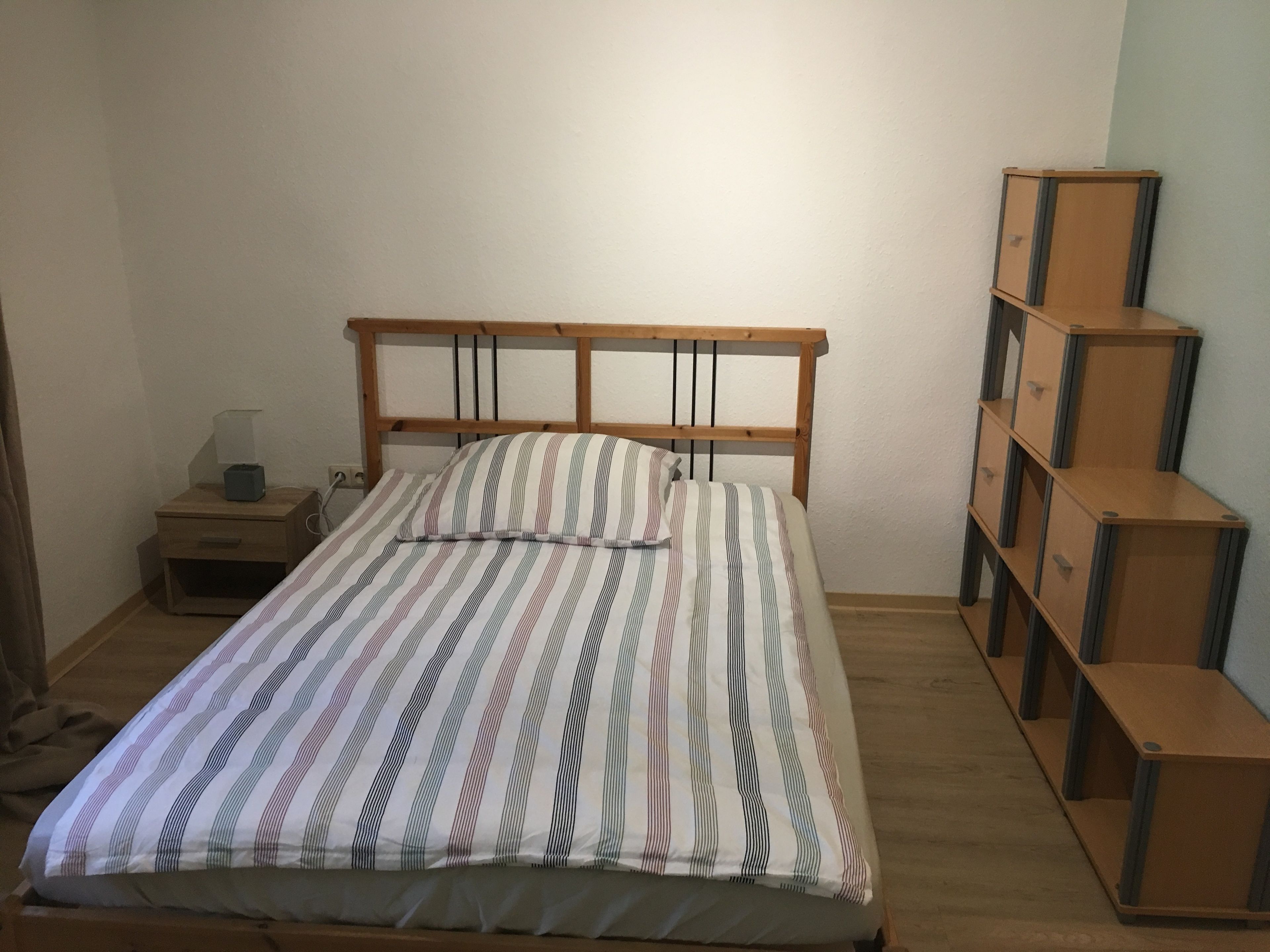 Quarto com estrutura de cama de madeira, mesa de cabeceira com abajur e armário embutido de madeira.