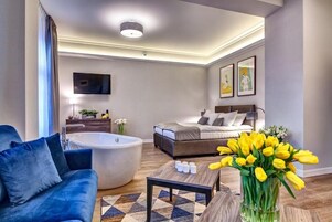 Quarto de hotel moderno com uma cama grande, área de estar com sofá e mesa de centro, banheira e televisão de tela plana fixada na parede.