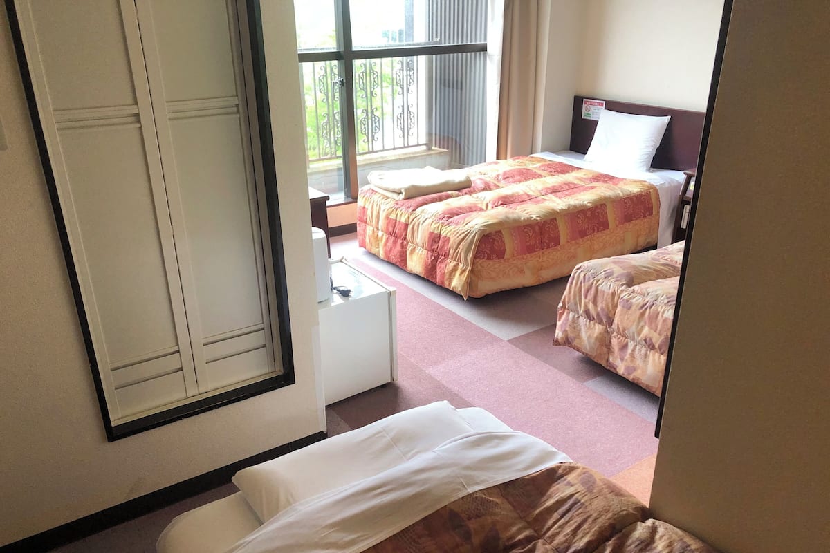 Quarto de hotel com duas camas, uma escrivaninha pequena e branca, e uma janela com vista para vegetação.