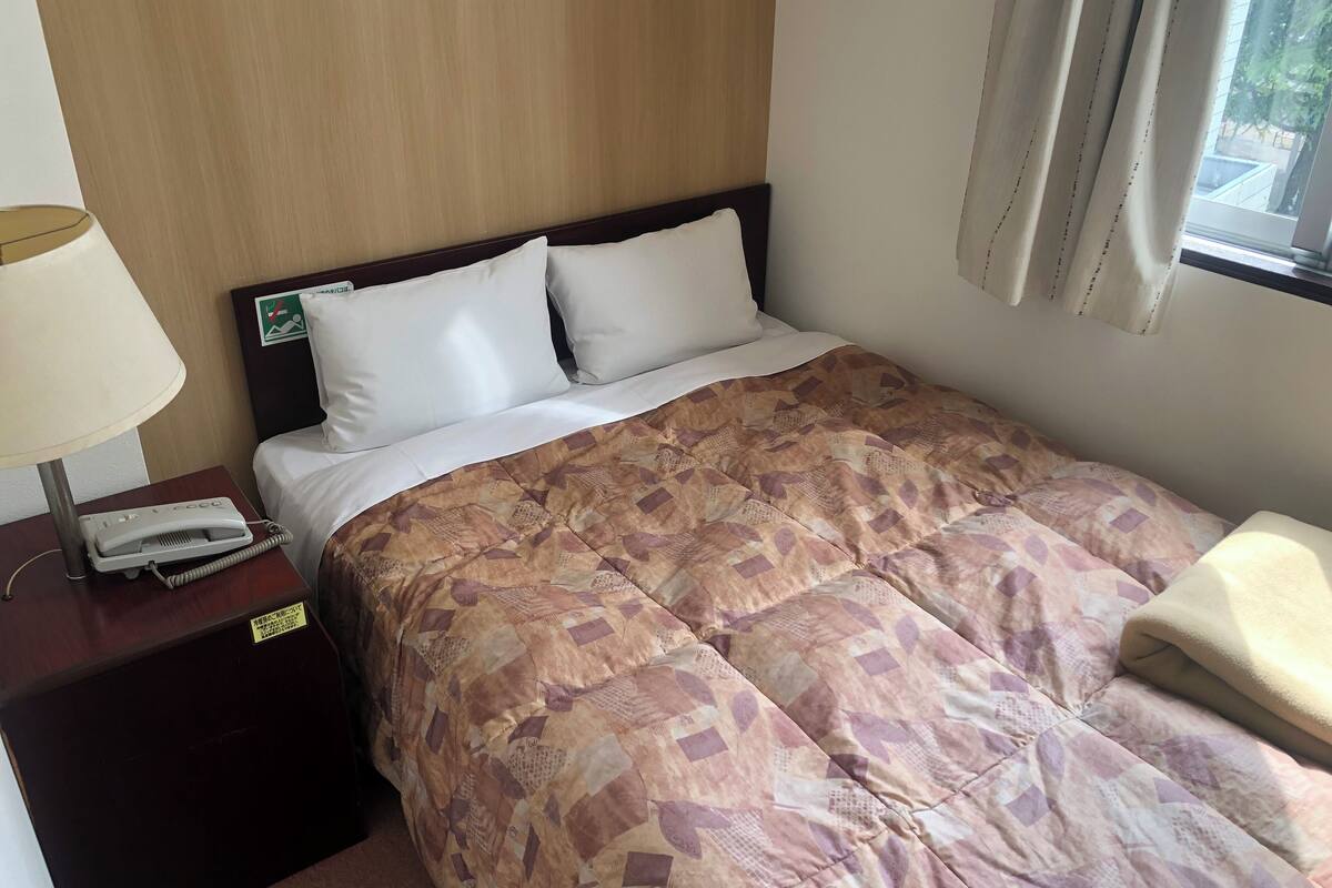 Quarto de hotel com uma cama, uma mesinha de cabeceira com abajur e telefone, e uma janela com cortinas.