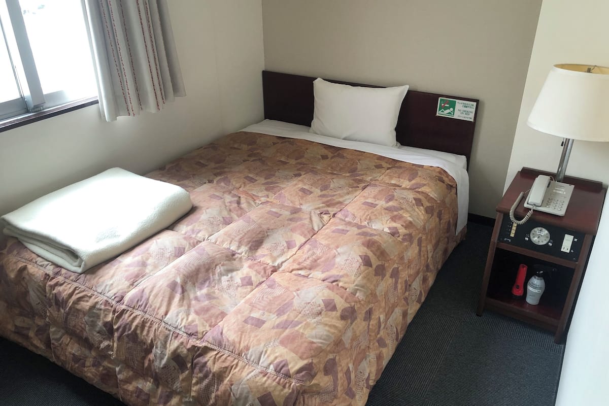 Quarto de hotel com cama, mesa de cabeceira com abajur e janela com cortinas.