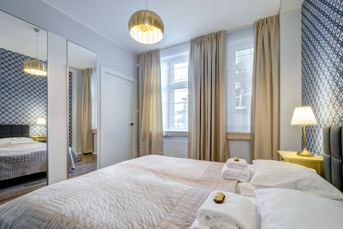 Quarto de hotel com uma cama grande, mesa de cabeceira com abajur, espelho e janela com cortinas.