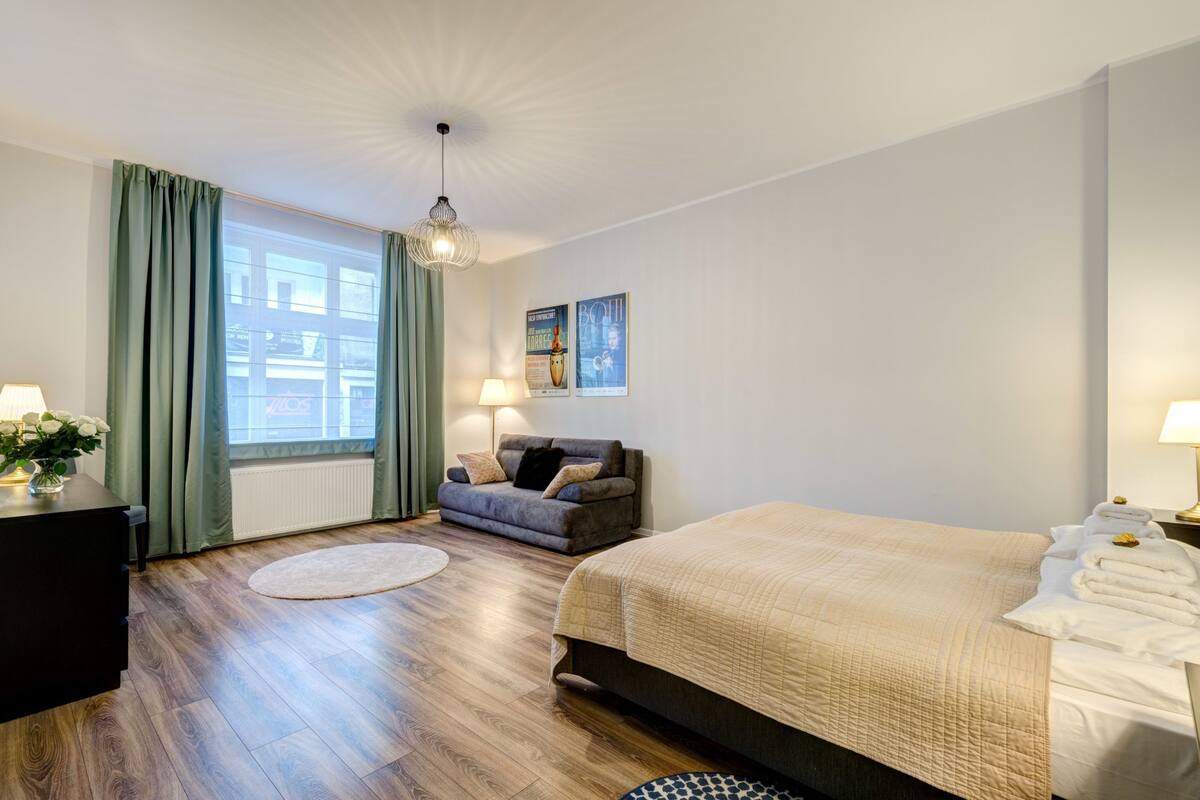 Quarto de hotel moderno com uma cama grande, um sofá, um tapete redondo, uma escrivaninha com abajur e vista para a cidade pela janela.