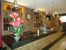Um bar de hotel com um balcão comprido, prateleiras com garrafas, um relógio e luminárias pendentes.