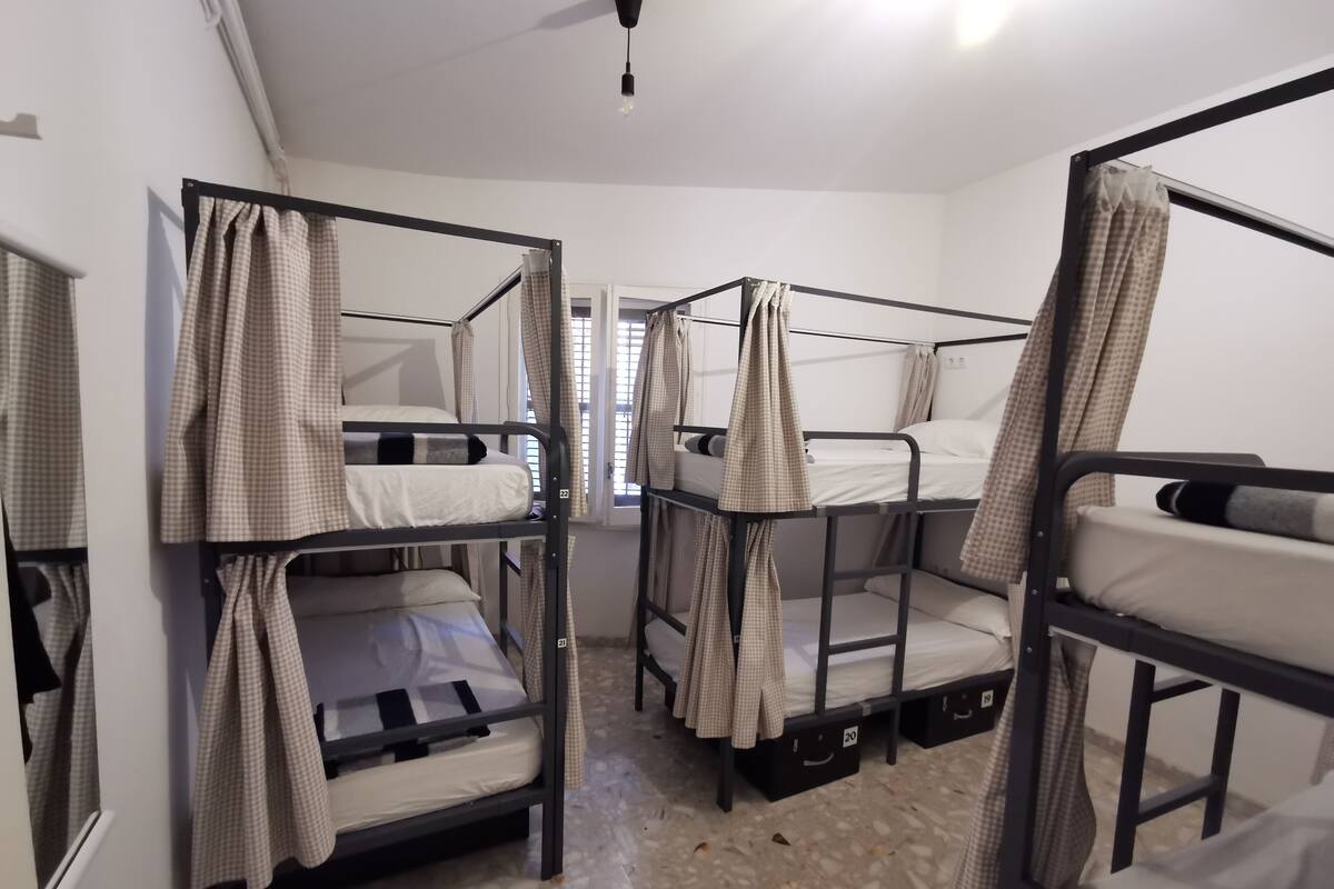 Quarto de dormitório com beliches, cada um com cortina. Há janelas com cortinas, uma luminária no teto e piso em azulejos.