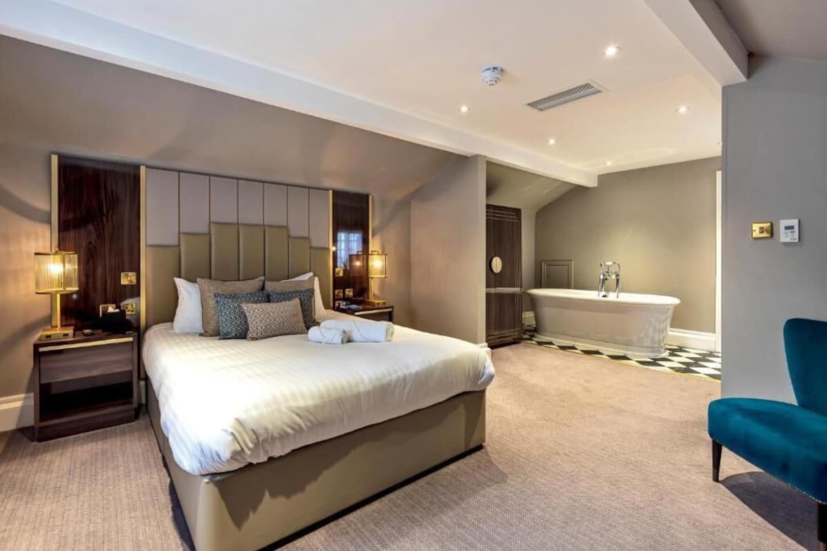 Quarto de hotel moderno com uma cama grande, área de estar com uma poltrona azul, banheira e banheiro separado.